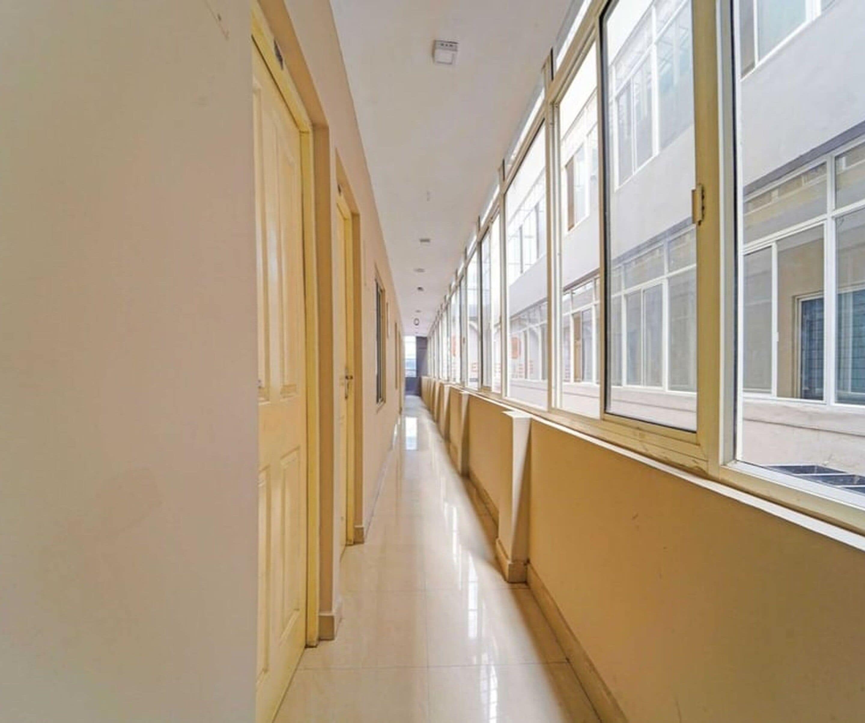 Um corredor comprido e estreito, com portas de um lado e janelas do outro, que dá acesso a um espaço interior iluminado.