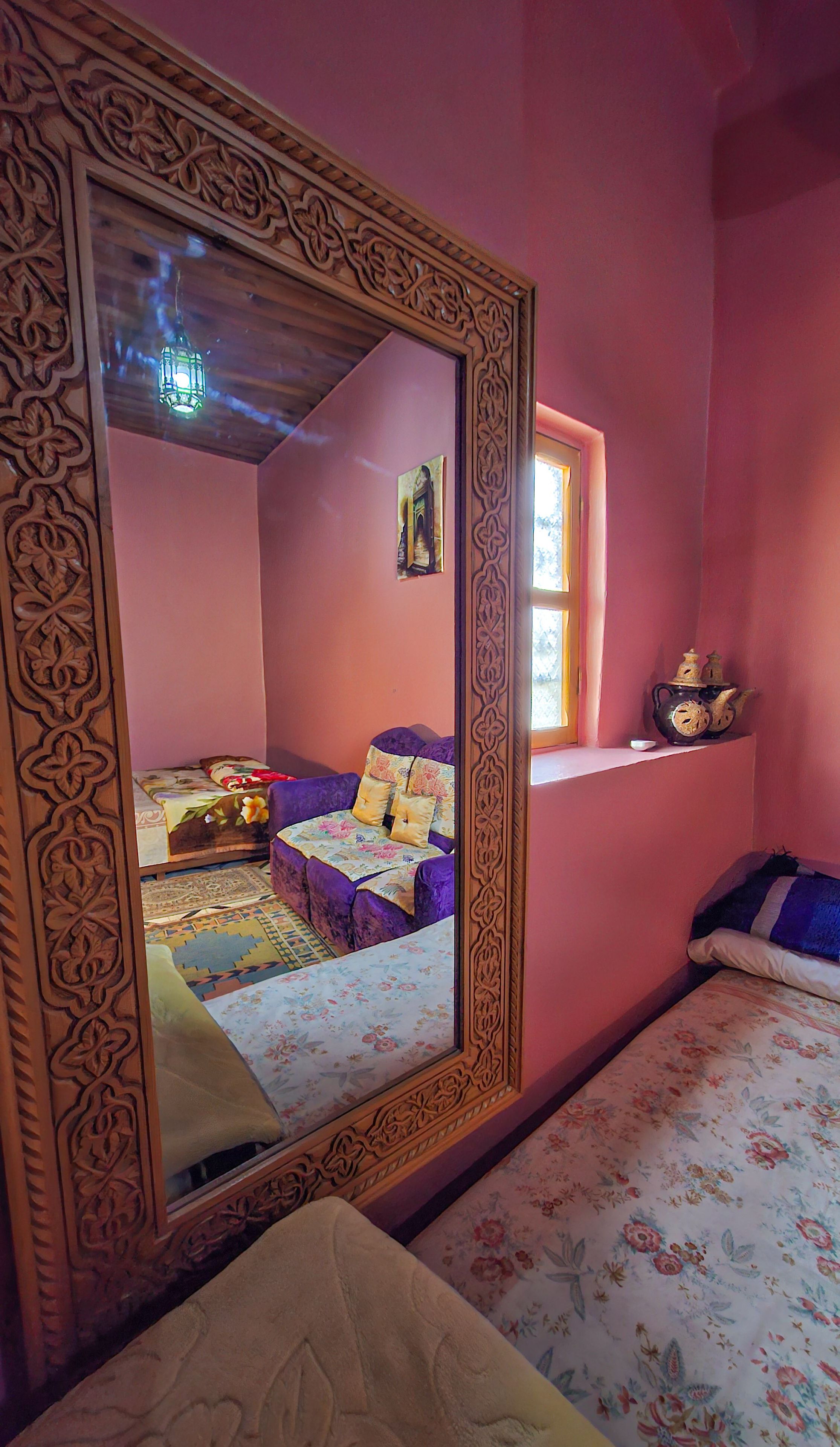 Quarto com um espelho grande e ornamentado, cama com roupa de cama floral, janela com vista e prateleira com objetos decorativos.