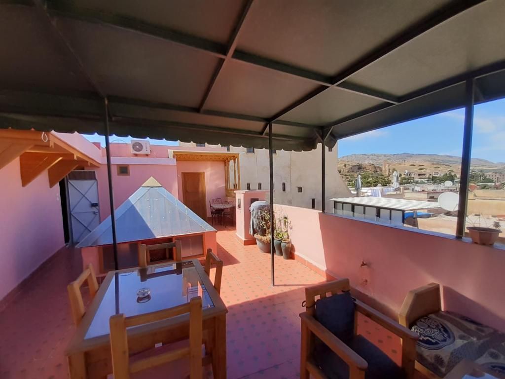 Uma área de estar coberta ao ar livre, com mesa e cadeiras, vista para uma paisagem montanhosa e um edifício com telhado plano ao fundo.