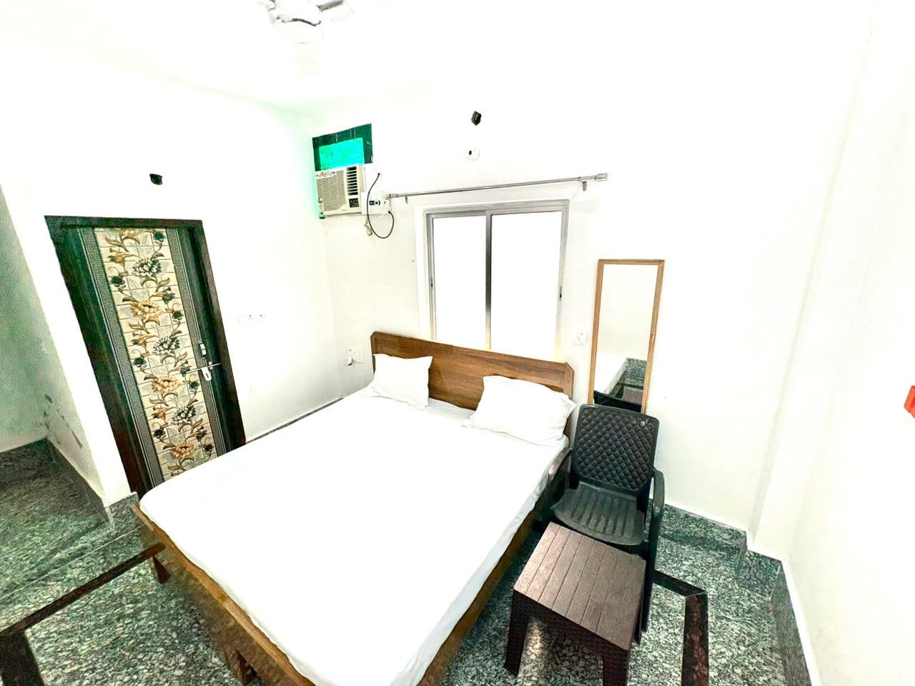 Quarto individual com uma cama e cabeceira de madeira, uma pequena mesa, uma cadeira, uma janela e um ar condicionado fixado na parede.