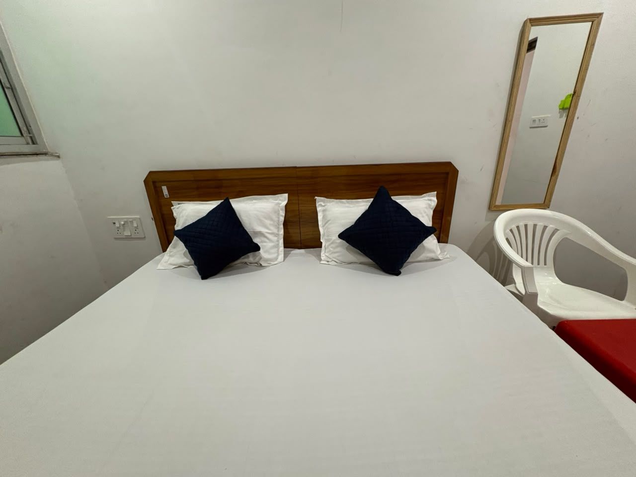 Uma cama de solteiro com cabeceira de madeira, duas almofadas, uma cadeira branca e um espelho na parede.