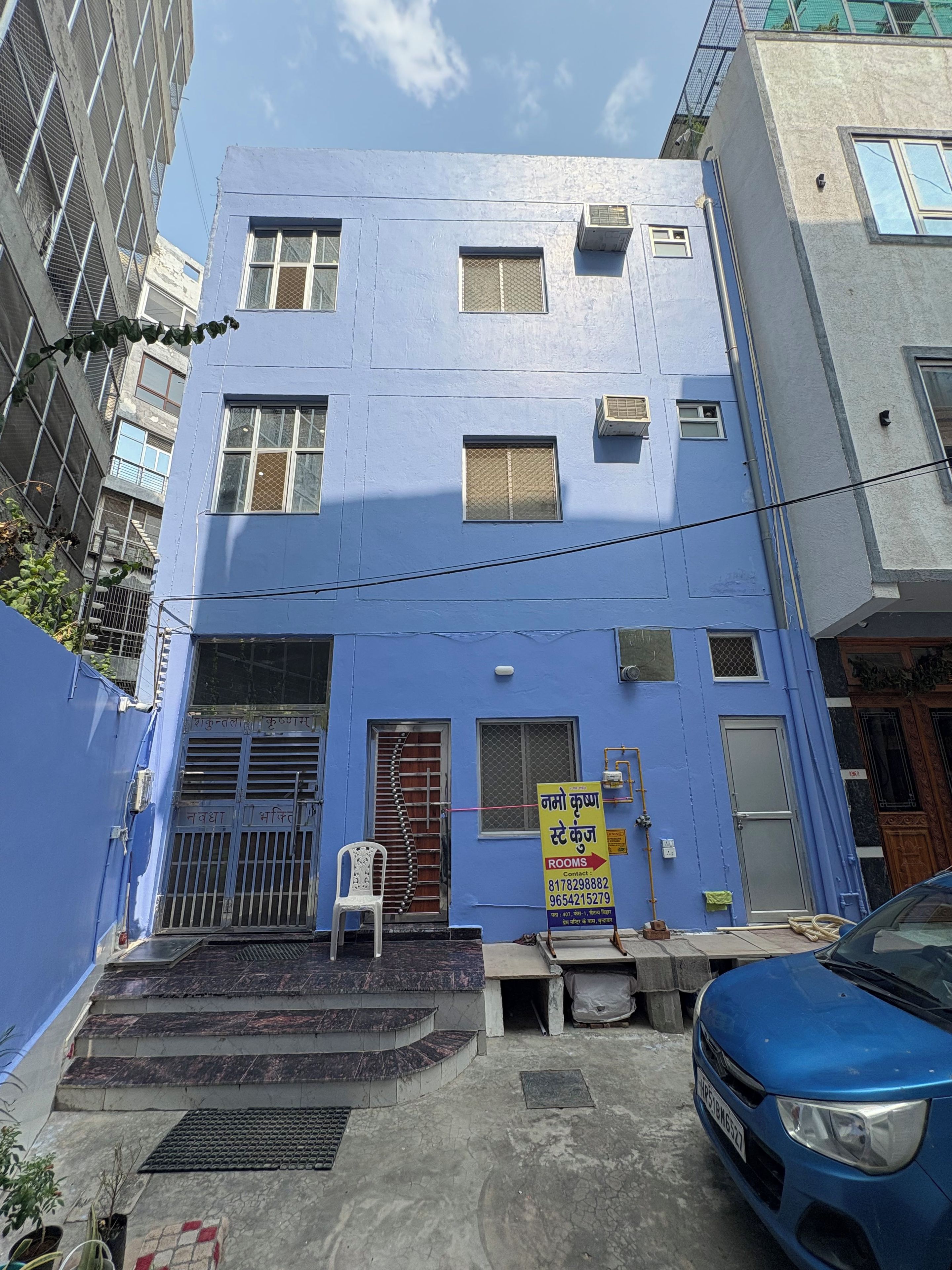 Um edifício de vários andares com fachada azul, uma escadaria visível para a entrada e um carro estacionado na frente.