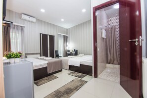 Quarto de hotel com duas camas, uma escrivaninha, uma cadeira, área de chuveiro e uma porta.