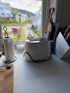 Uma bancada de cozinha com uma torradeira, uma tábua de corte e uma janela com vista para casas e uma estrada.