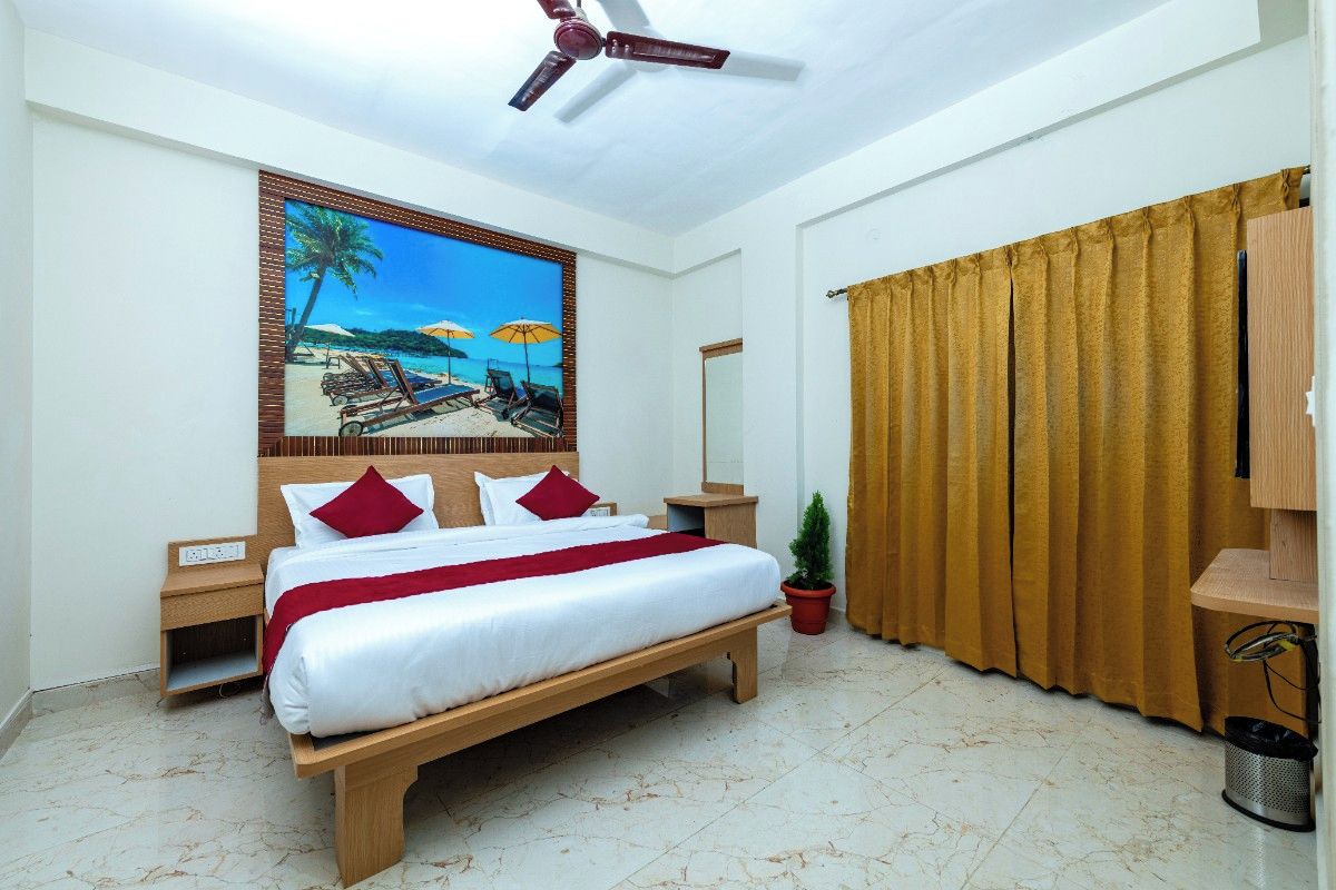 Quarto de hotel com cama, ventoinha no teto, pintura de uma praia, mesa de cabeceira, planta em vaso e janela com cortinas.