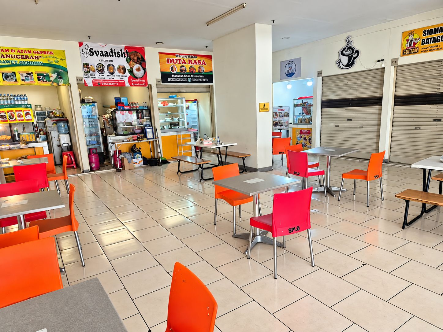 Uma praça de alimentação com várias áreas de estar, mesas e cadeiras, e diversos quiosques de comida com placas visíveis.