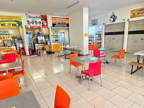 Uma praça de alimentação com várias áreas de estar, mesas e cadeiras, e diversos quiosques de comida com placas visíveis.
