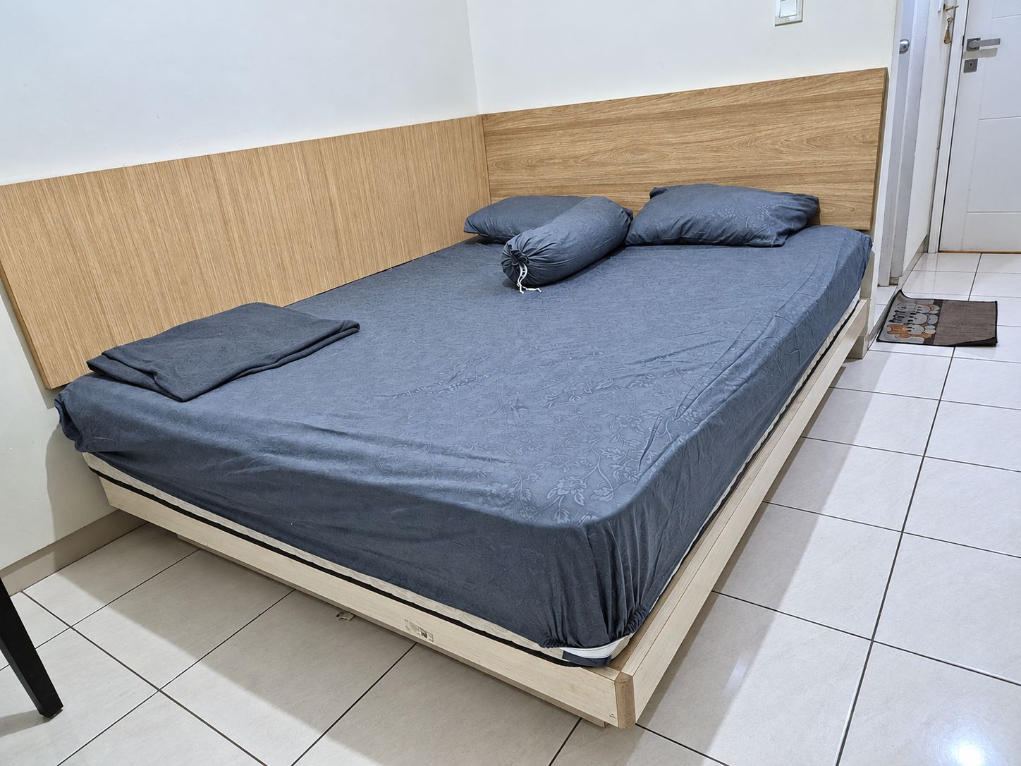 Uma cama de solteiro com cabeceira de madeira, colcha cinza escura e almofadas a combinar.