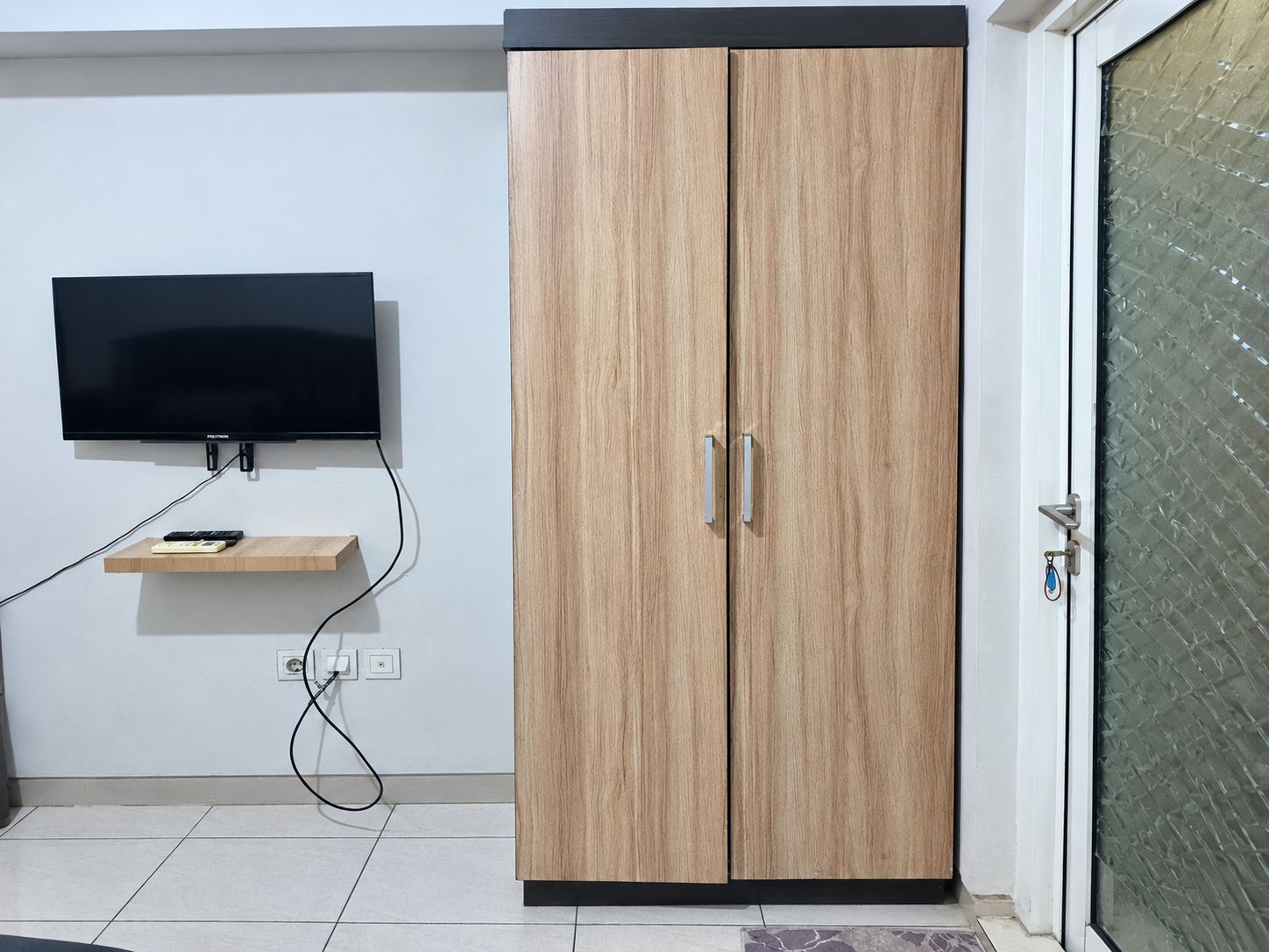 Quarto com guarda-roupa de madeira, televisão montada na parede e porta com painel de vidro texturizado.