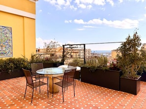 Terraço com mesa e cadeiras de vidro, rodeado por plantas em vasos e chão de azulejos.