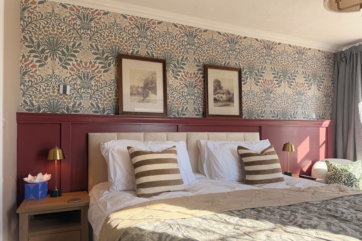Quarto de hotel com uma cama grande, duas fotografias emolduradas na parede, uma mesinha de cabeceira com abajur e um aparador de madeira.