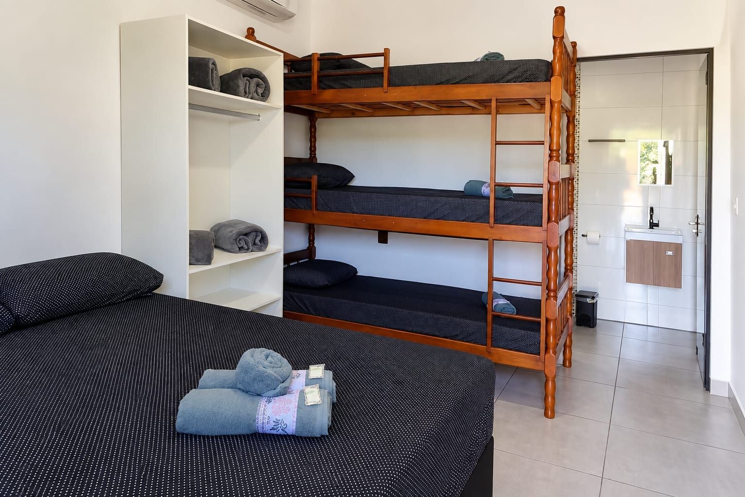 Quarto com beliche, guarda-roupa branco, banheiro pequeno e cama com colcha estampada.