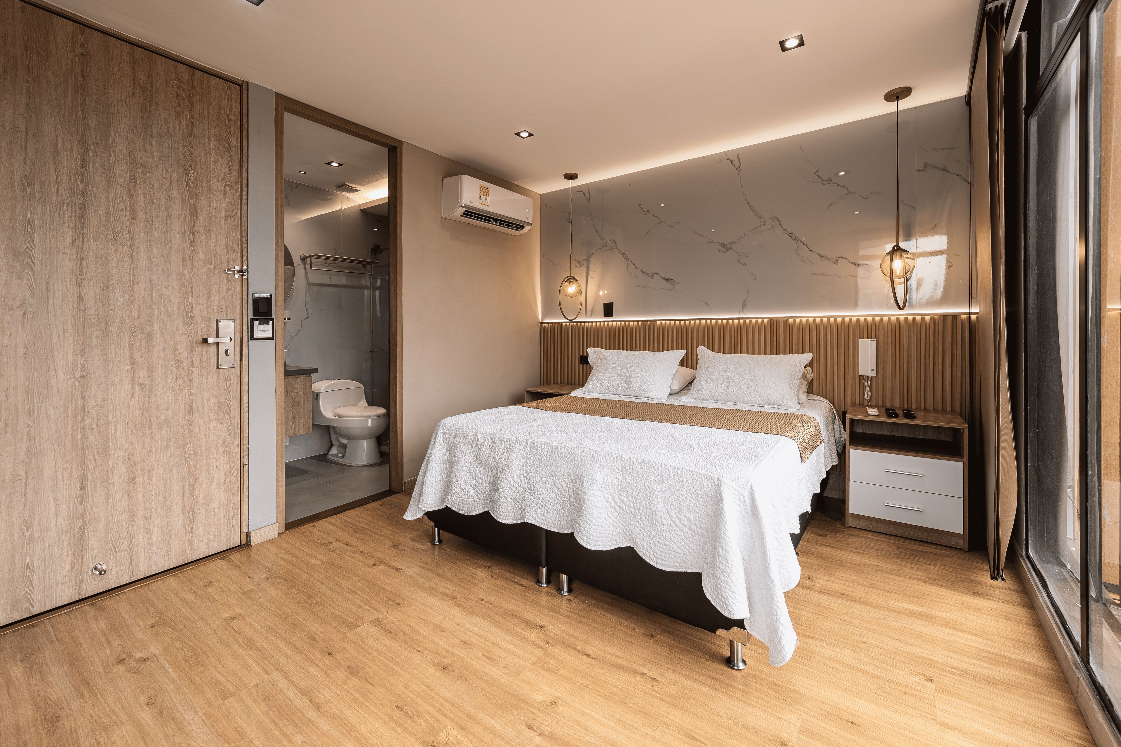 Quarto moderno com piso de madeira, cama com colcha branca, mesa de cabeceira com abajur e um espelho grande na parede.