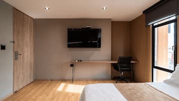 Quarto moderno com piso de madeira, cama com manta estampada, escrivaninha com cadeira, TV na parede e janela com persianas.