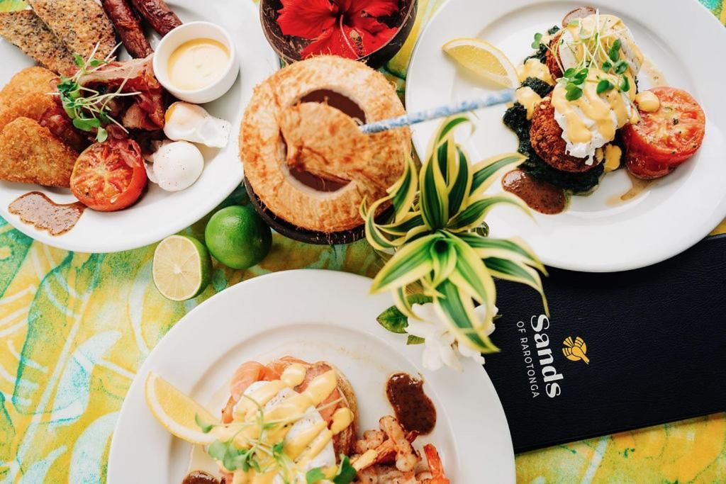 Diversos pratos de café da manhã em placas, incluindo ovos, bacon e torradas, com uma planta tropical e uma toalha de mesa preta com o logo 'Sands of Rarotonga' ao fundo.