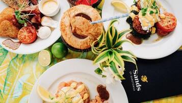 Diversos pratos de café da manhã em placas, incluindo ovos, bacon e torradas, com uma planta tropical e uma toalha de mesa preta com o logo 'Sands of Rarotonga' ao fundo.