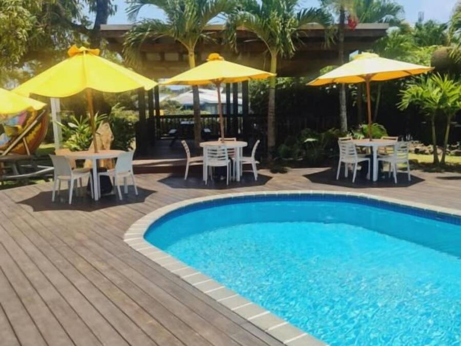 Piscina com deck de madeira, rodeada por mesas e cadeiras sob guarda-sóis amarelos, com palmeiras ao fundo.