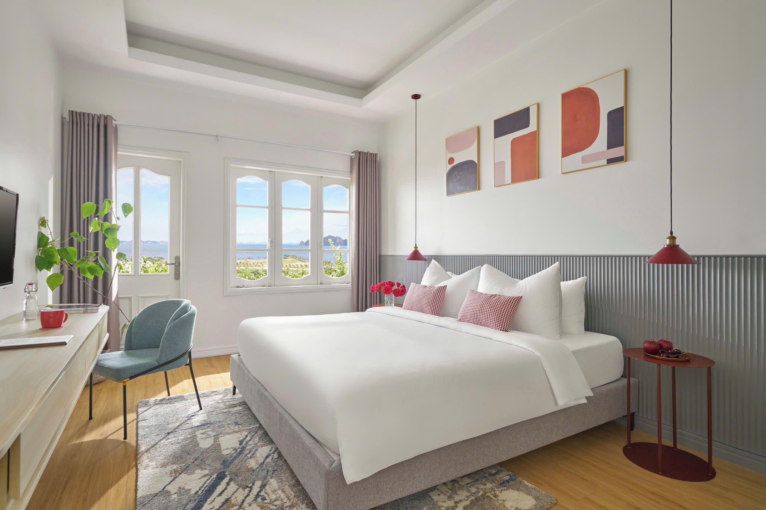 Quarto de hotel moderno com cama grande, escrivaninha, cadeira e vista para o mar.