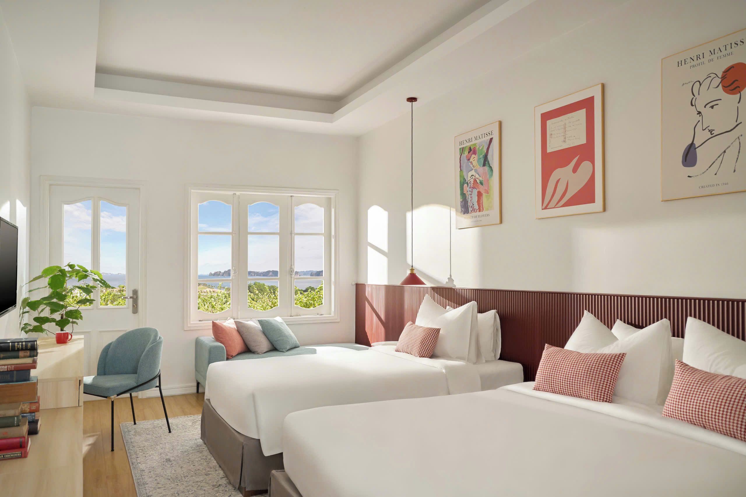 Quarto de hotel com duas camas, uma escrivaninha, uma cadeira e uma janela grande com vista.