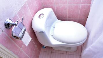 Sanitário com vaso sanitário branco, função bidê e dispensador de toalhas de papel, em um banheiro com paredes revestidas de azulejos rosa.