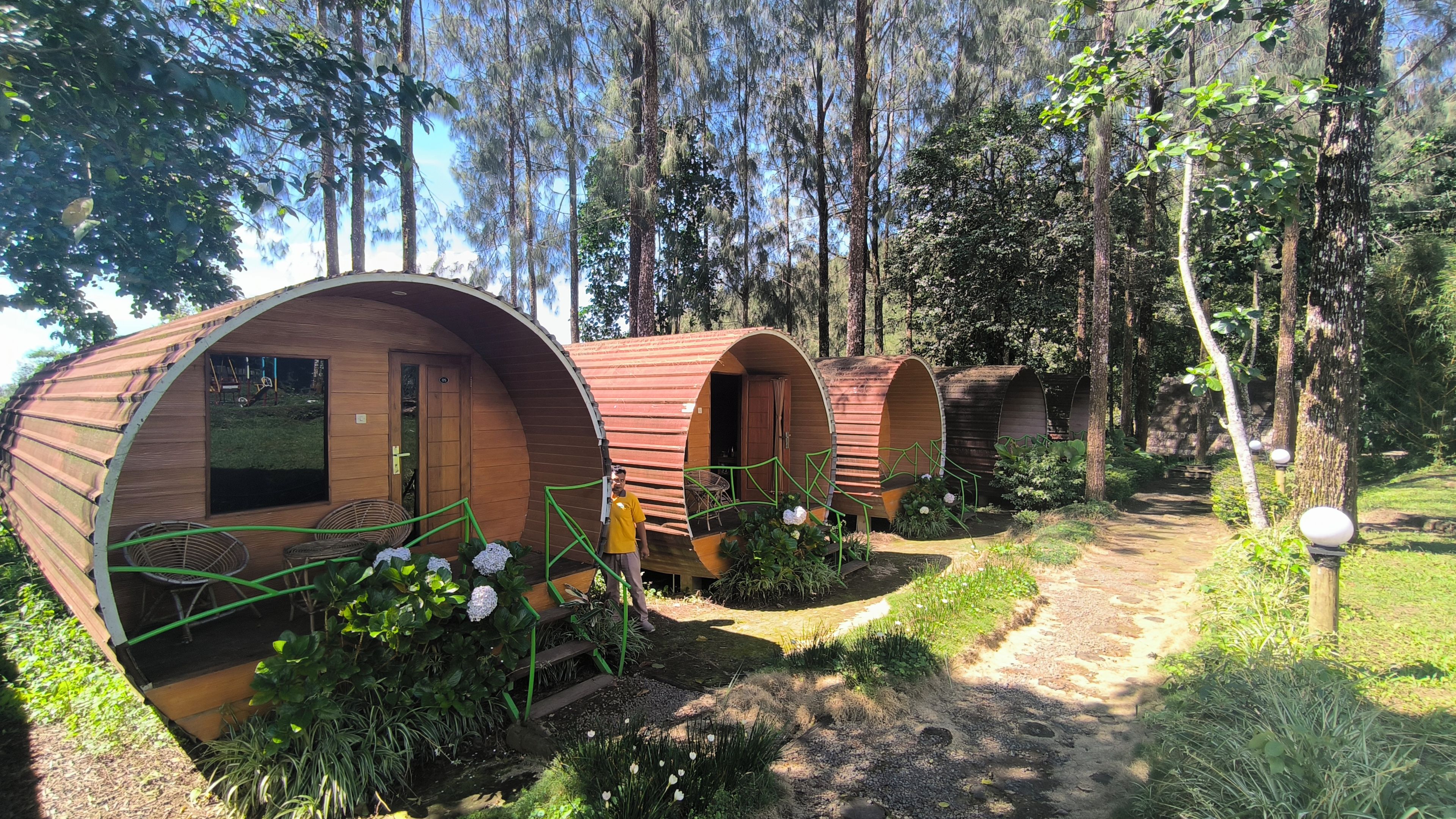 Fileira de cabanas em formato de barril de madeira, rodeada por área arborizada com trilha de terra passando por elas.