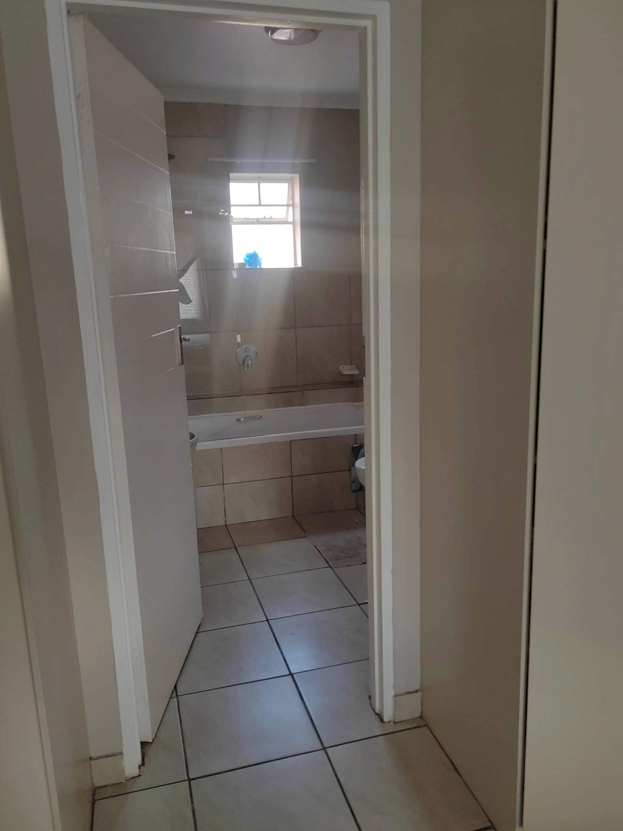 Corredor estreito que dá para um banheiro com janela, pia e vaso sanitário.