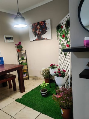 Uma área de refeições com mesa de madeira, cadeiras, luminária pendente, quadro na parede, plantas em vasos e tapete de grama artificial verde.
