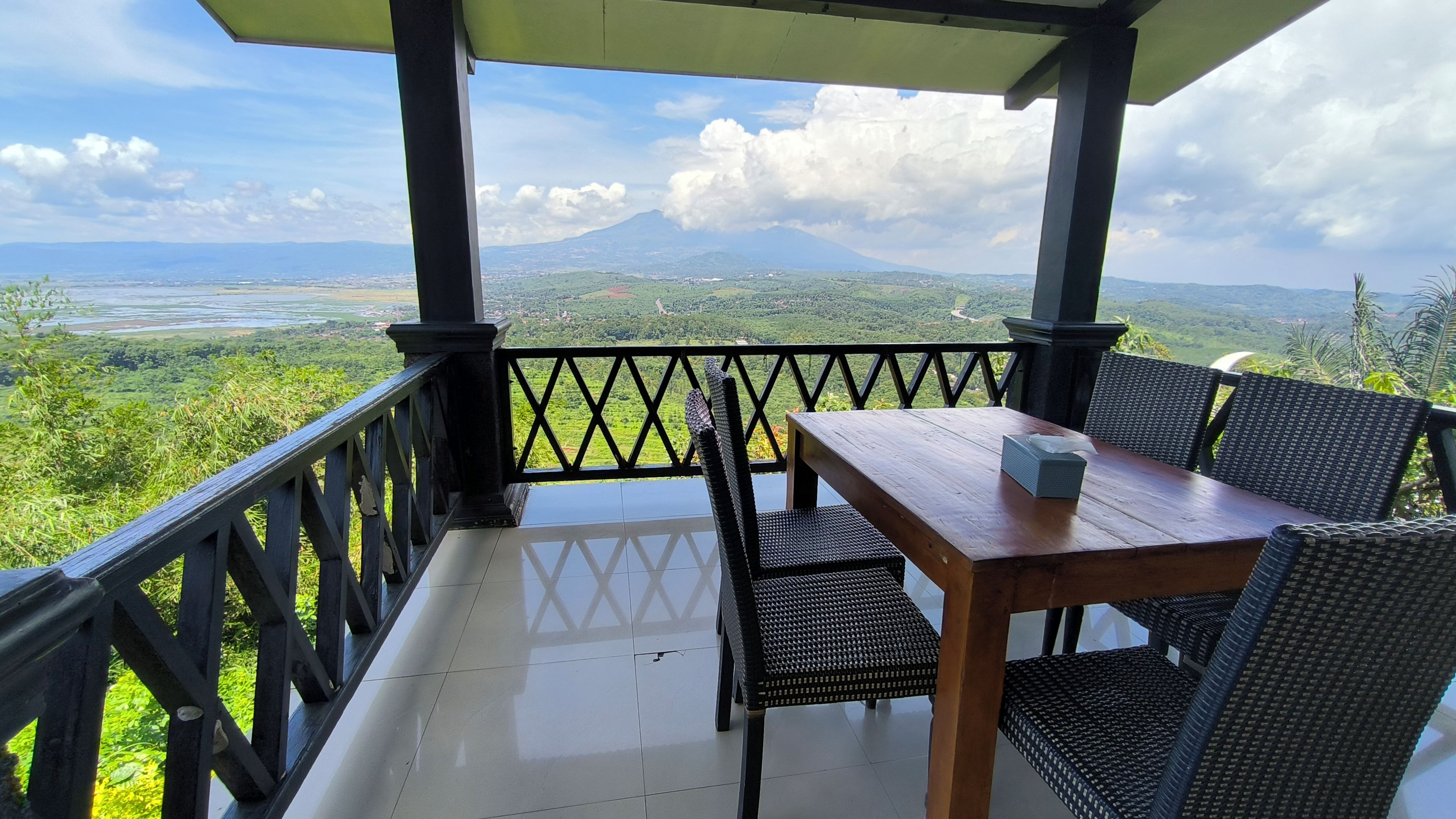 Uma área de estar coberta ao ar livre, com mesa e cadeiras, com vista para uma paisagem exuberante e uma montanha ao longe.