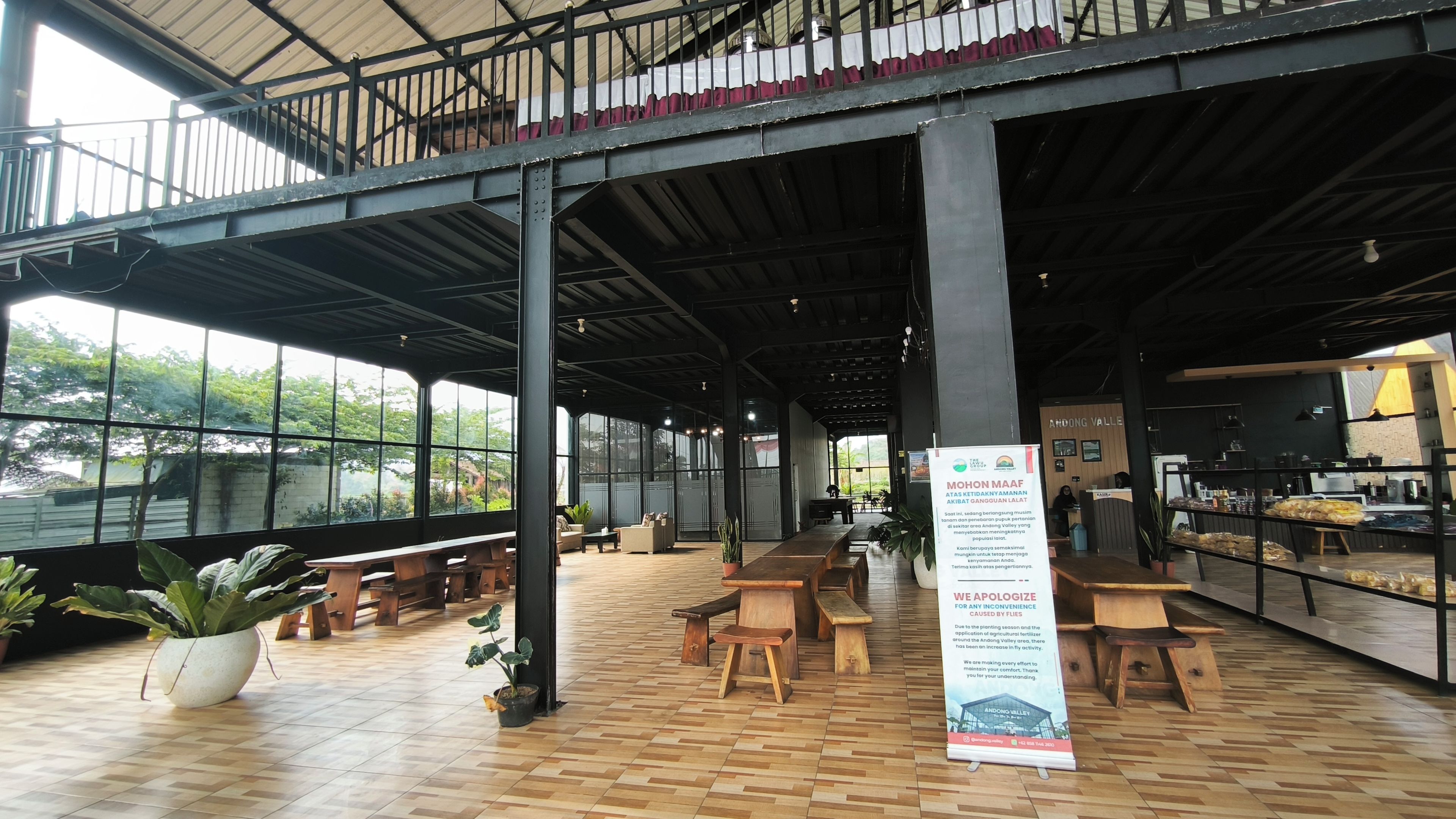 Espaço interno com bancos e mesas de madeira, janelas amplas e uma placa.