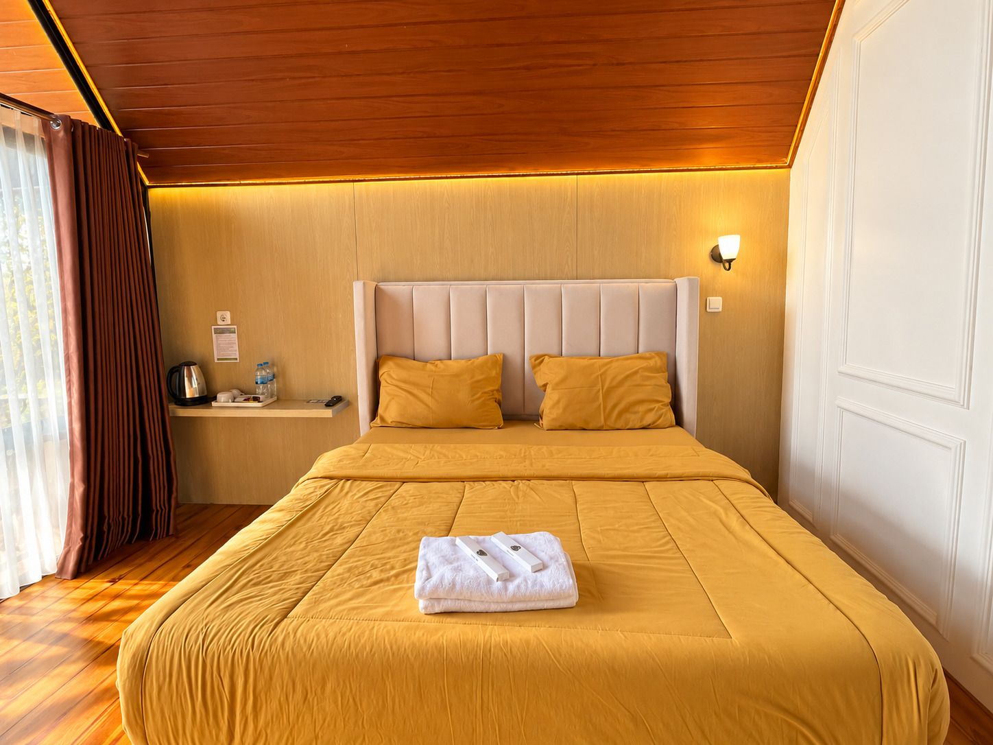 Quarto de hotel com cama, mesa de cabeceira, luminária na parede e janela com cortinas.