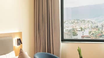 Quarto de hotel com cama, cadeira, mesinha, mesa de cabeceira e janela com vista para a cidade.