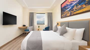 Quarto de hotel com cama, escrivaninha, cadeira, televisão, janela com cortinas e um quadro na parede.