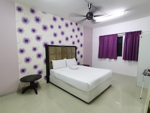 Quarto com cama, ventilador de teto, mesinha e janela com cortinas roxas.