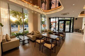 Um interior moderno com sofá, mesas de jantar e cadeiras, janelas amplas com cortinas e vista para a rua.