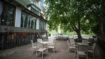 Uma área externa com mesas e cadeiras de vime, um edifício com grandes janelas e uma árvore que oferece sombra.