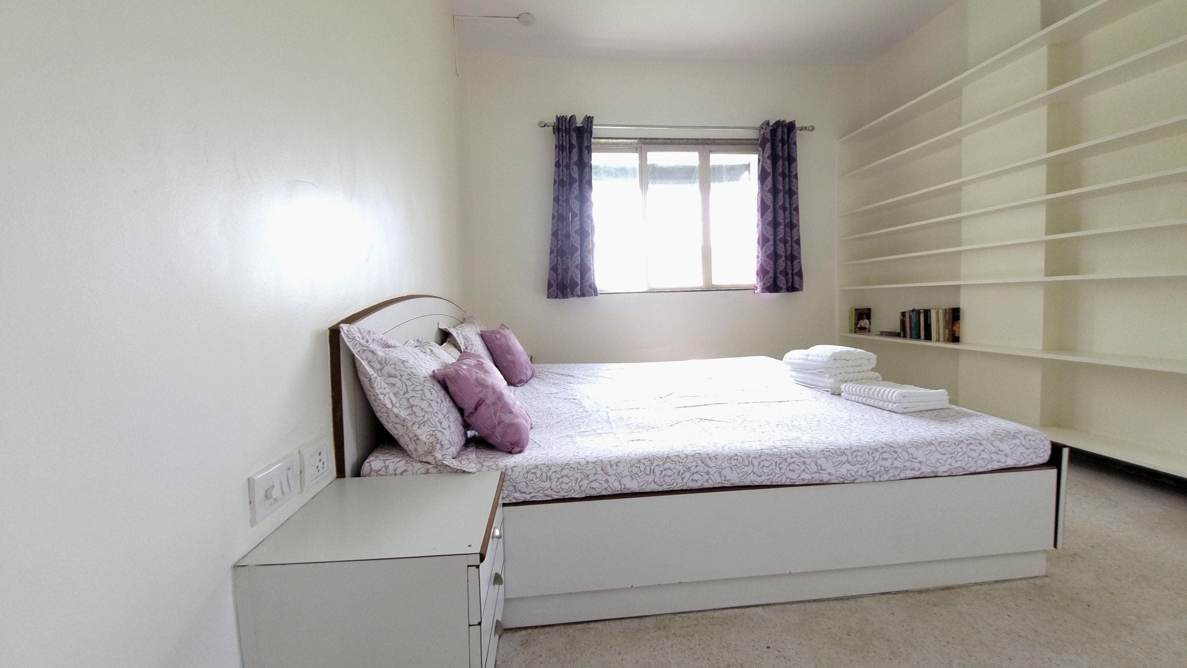 Quarto com cama, mesa de cabeceira, janela com cortinas e guarda-roupa embutido.