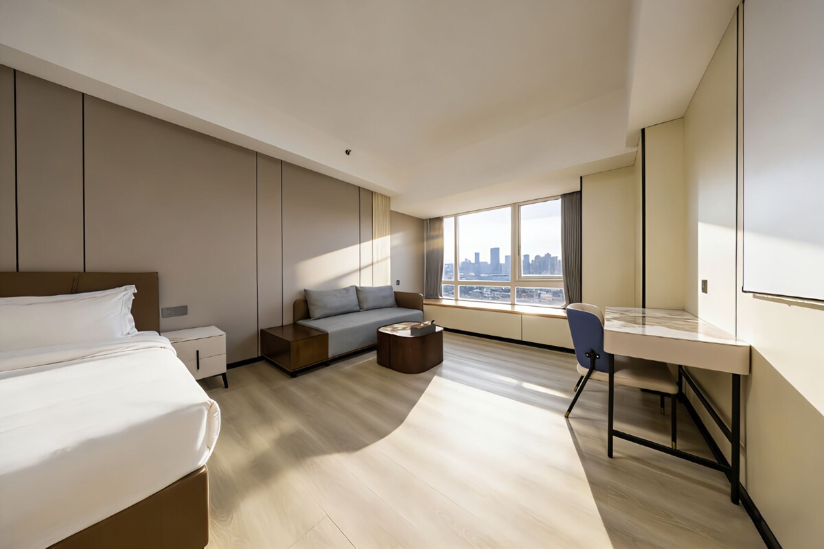 Quarto de hotel moderno com cama, sofá, escrivaninha e uma grande janela com vista para a cidade.