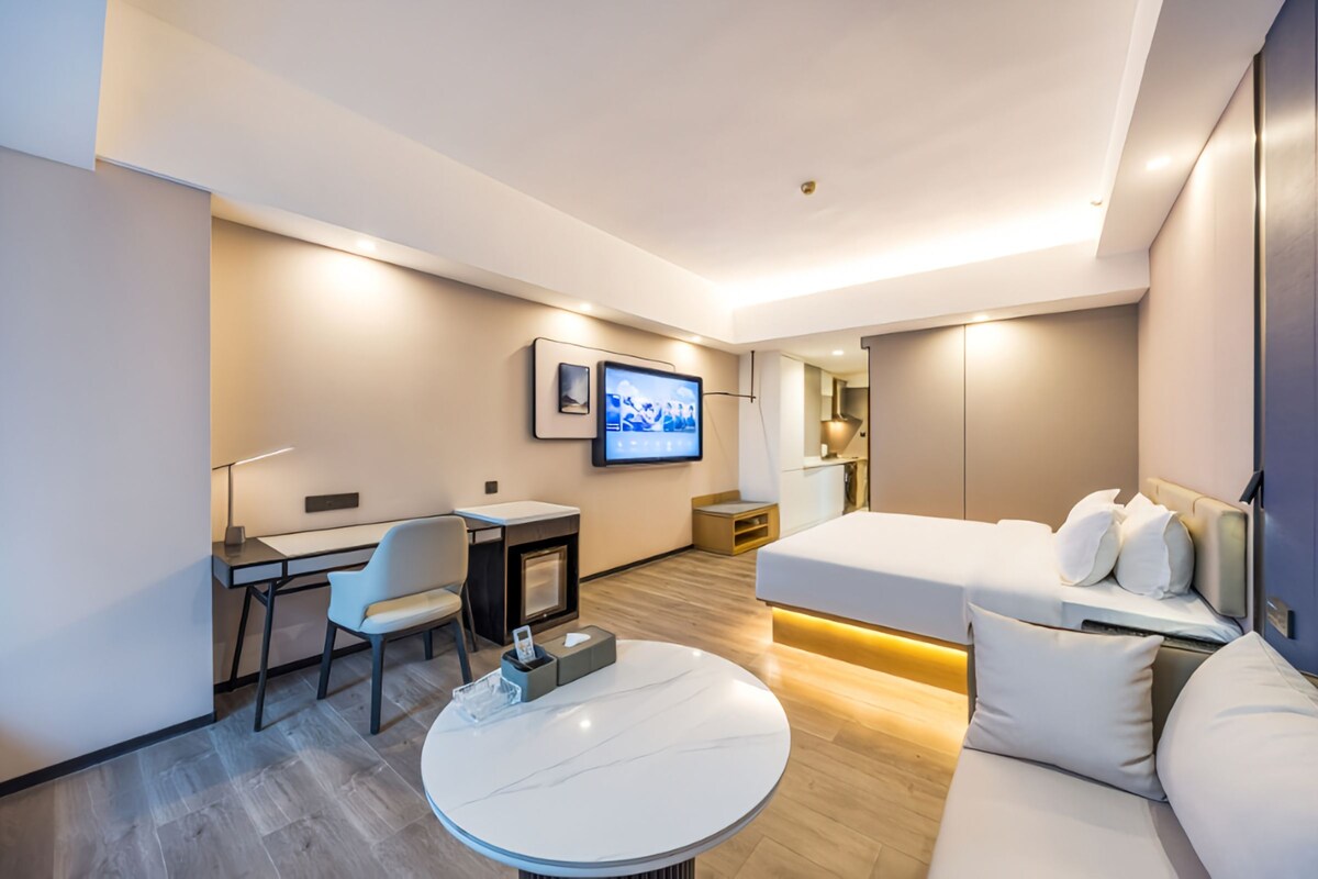 Quarto de hotel moderno com cama, escrivaninha, cadeira, mesa redonda e TV fixada na parede.