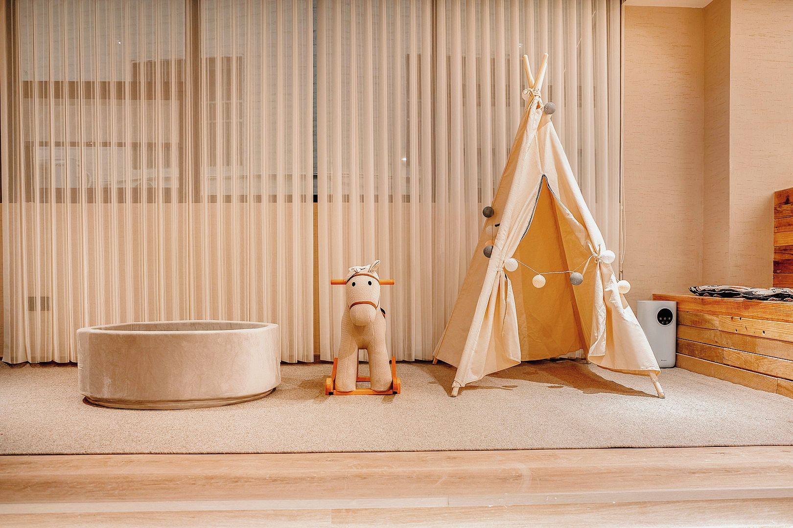 Quarto com uma janela grande, piso de madeira, cama redonda, um pequeno brinquedo de cavalo de madeira e um tipi branco.