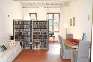 Uma sala de estar aconchegante com um sofá, uma mesa de jantar com cadeiras, uma estante de livros e uma janela com cortinas.