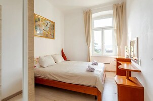 Quarto com cama, mesa de cabeceira de madeira, janela com cortinas e um quadro na parede.