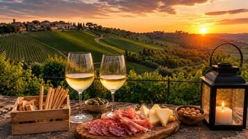 Vista panorâmica de um vinhedo, com uma mesa posta para refeição, incluindo palitos de pão, queijos, embutidos, azeitonas e copos de vinho branco.