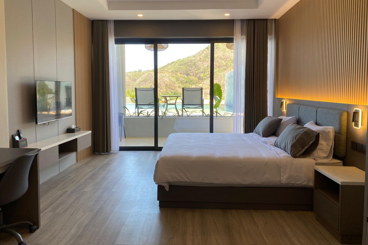 Quarto de hotel moderno com uma cama grande, escrivaninha, televisão e varanda com vista para o exterior.