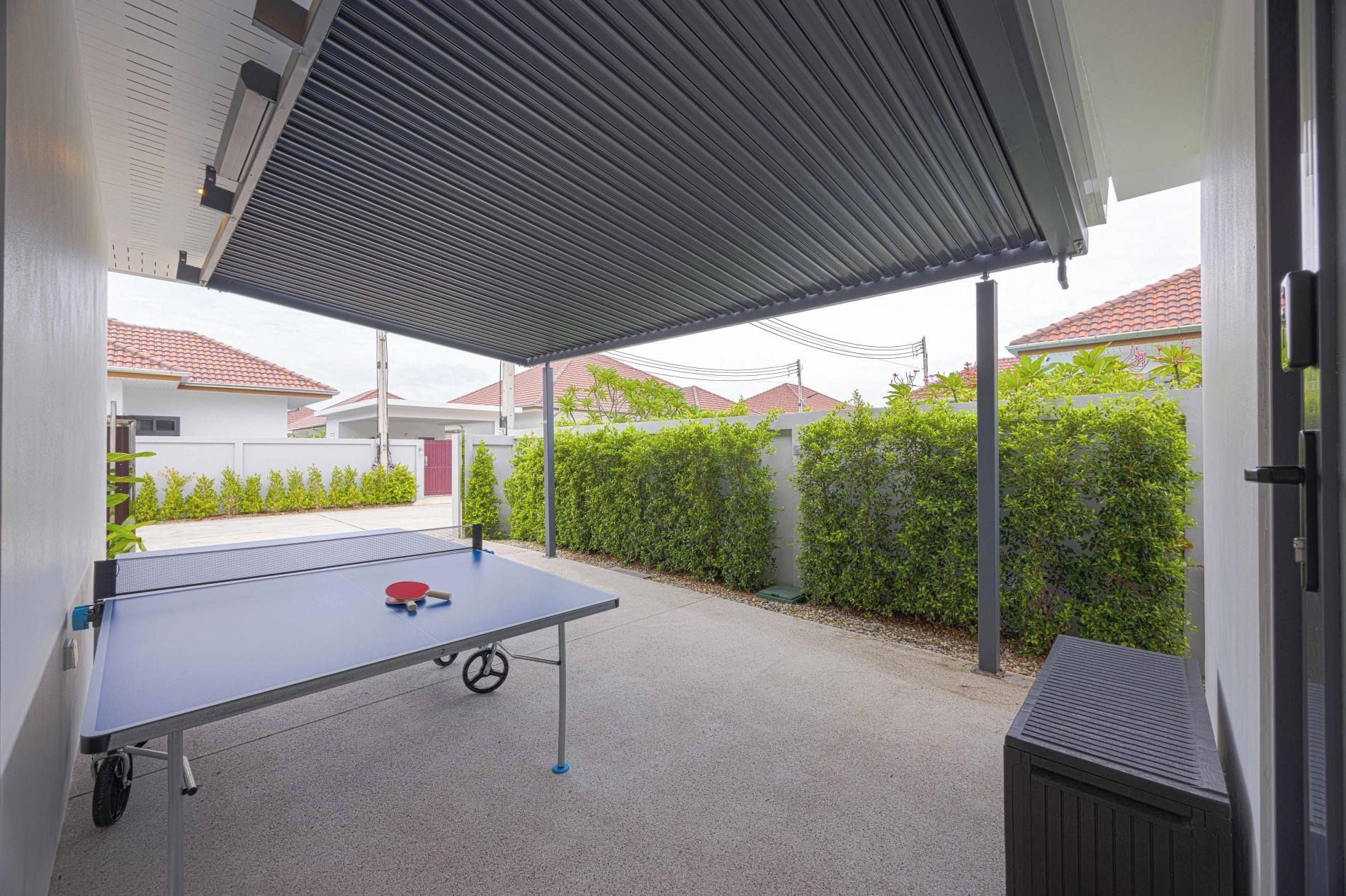 Mesa de pingue-pongue ao ar livre, sob uma cobertura, com vista para um bairro residencial.