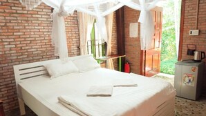 Uma cama com dossel, uma mesa pequena com uma cadeira, um refrigerador e uma parede de tijolos.