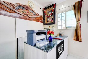 Área de recepção de hotel com balcão, relógio, quadro emoldurado, vaso com flores e janela com cortinas.