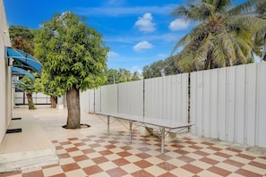 Um pátio externo com piso xadrez, uma mesa de metal e um cercado branco.