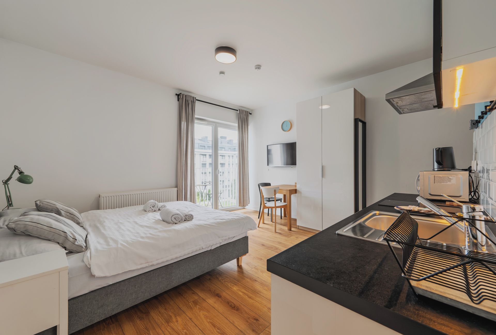Quarto de hotel moderno com cama, escrivaninha, cadeira, televisão e cozinha compacta.