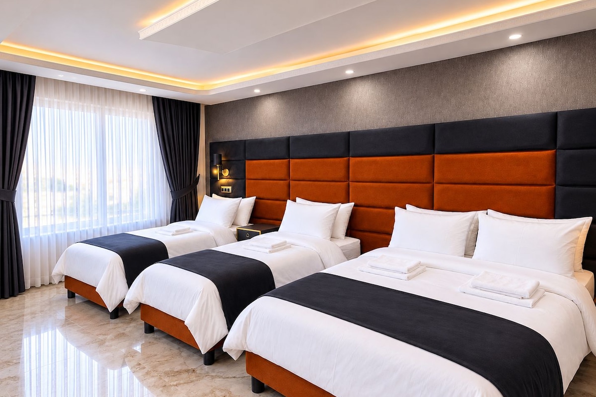 Quarto de hotel com três camas, uma janela grande com cortinas e uma cabeceira moderna.