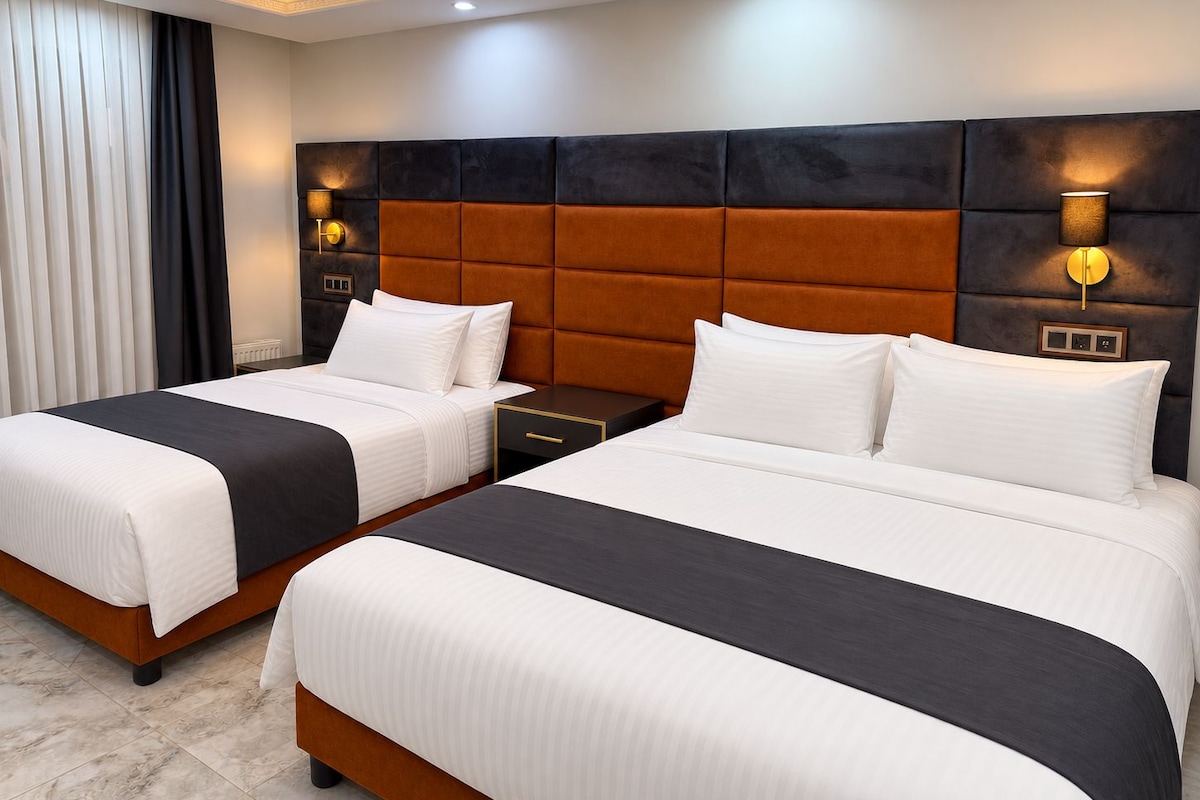Quarto de hotel com duas camas, cada uma com roupa de cama branca e cabeceira escura. Há mesas de cabeceira com luminárias, e o quarto tem um design moderno com piso de mármore.